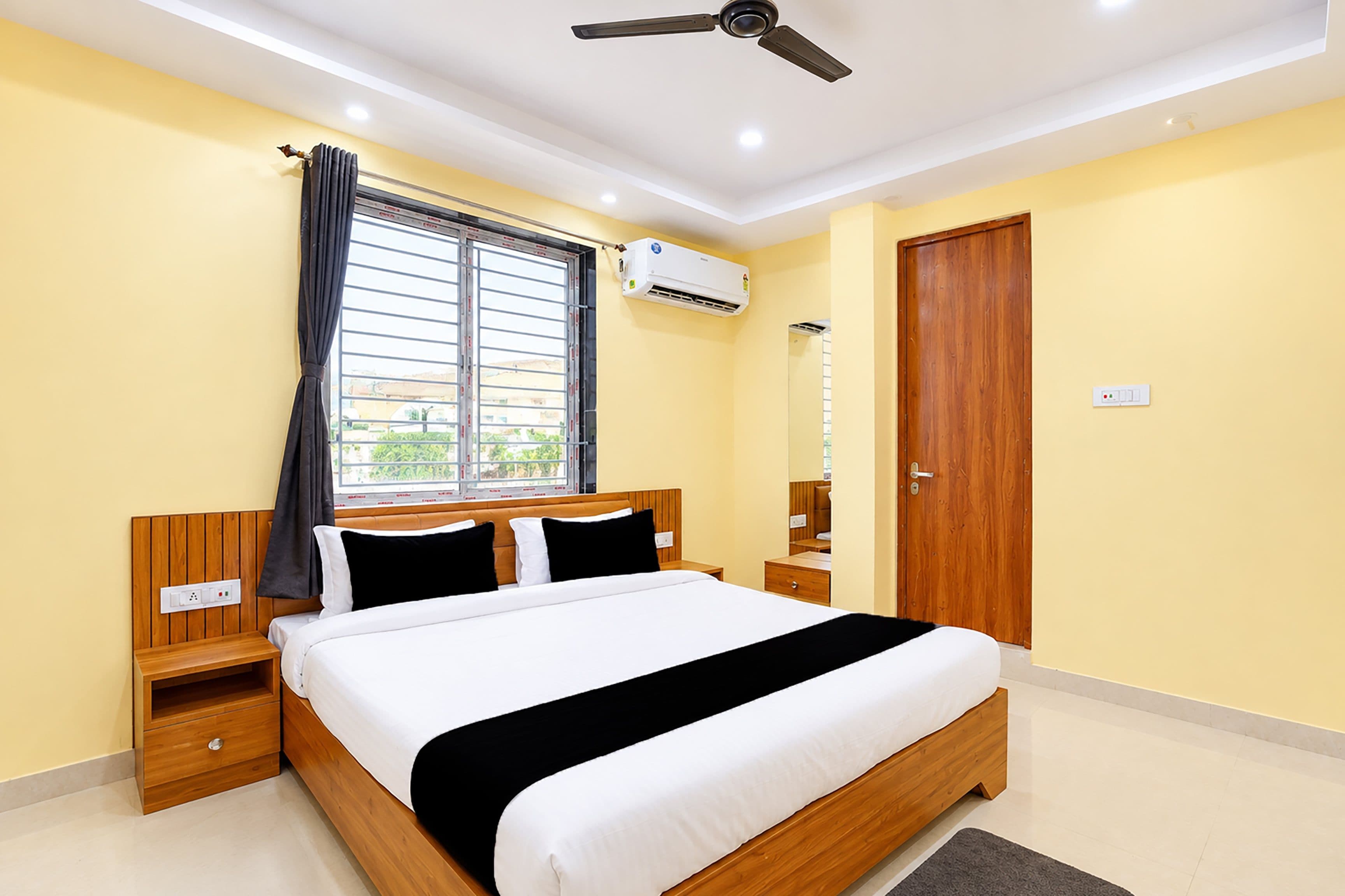 Quarto com cama, mesas de cabeceira, ventoinha no teto, ar condicionado, janela com cortinas e porta de madeira.