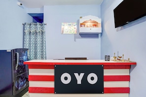 Balcão de recepção com uma placa indicando 'OYO' em um cômodo com paredes azul claro, uma televisão montada e um pequeno altar com objetos religiosos.