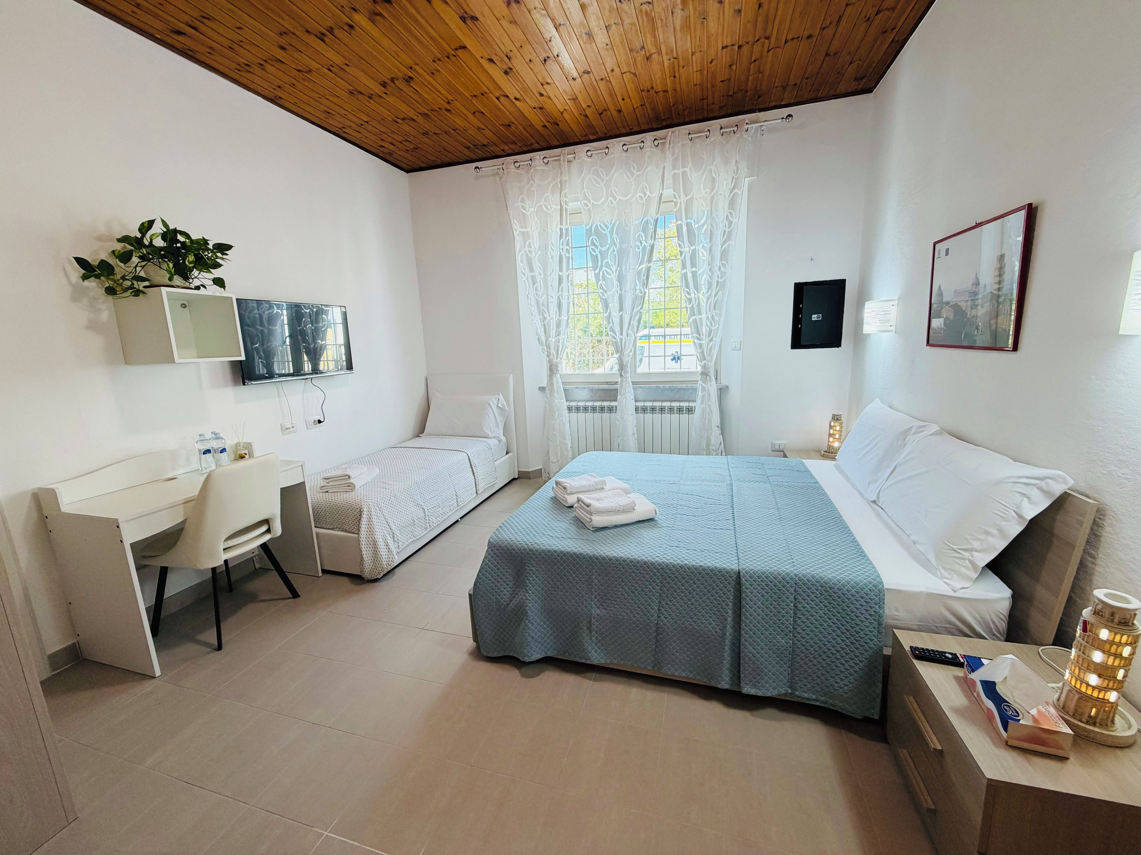Quarto de hotel com cama, escrivaninha, cadeira, televisão, janela com cortinas e mesa de cabeceira com abajur.