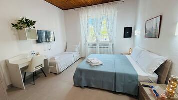 Quarto de hotel com cama, escrivaninha, cadeira, televisão, janela com cortinas e mesa de cabeceira com abajur.