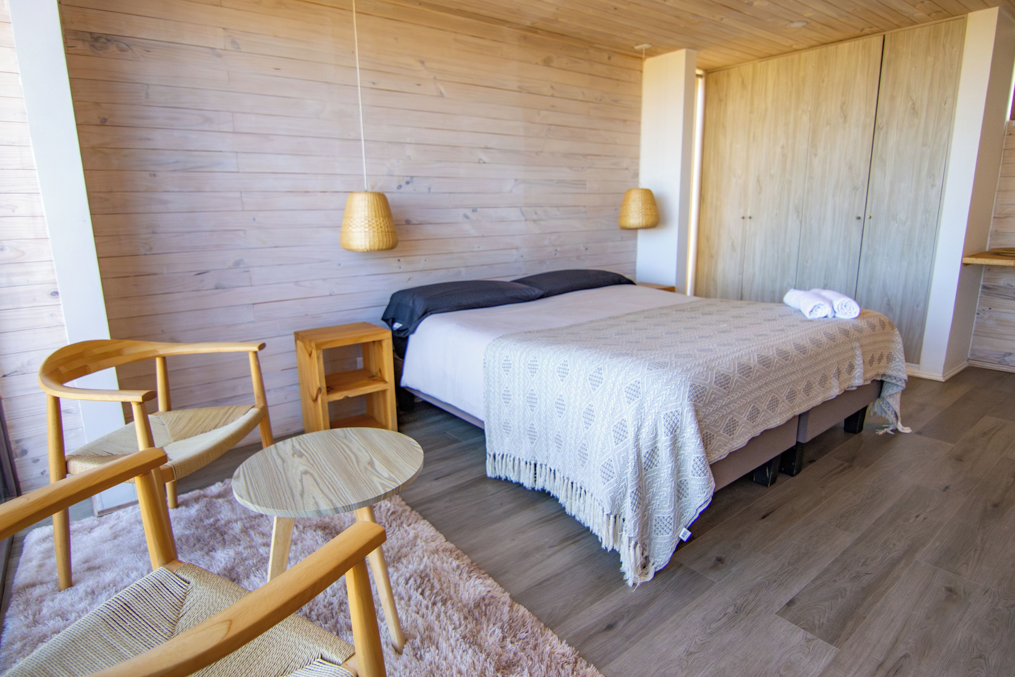 Quarto com cama, duas cadeiras, uma mesinha e chão de madeira.