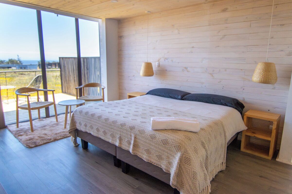 Quarto com uma cama grande, piso de madeira, uma mesa pequena, duas cadeiras e vista por uma porta de vidro.