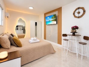 Quarto de hotel moderno com cama, televisão, área de refeições com cadeiras e relógio fixado na parede.