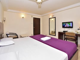 Quarto de hotel com cama, escrivaninha, cadeira, televisão e espelho.