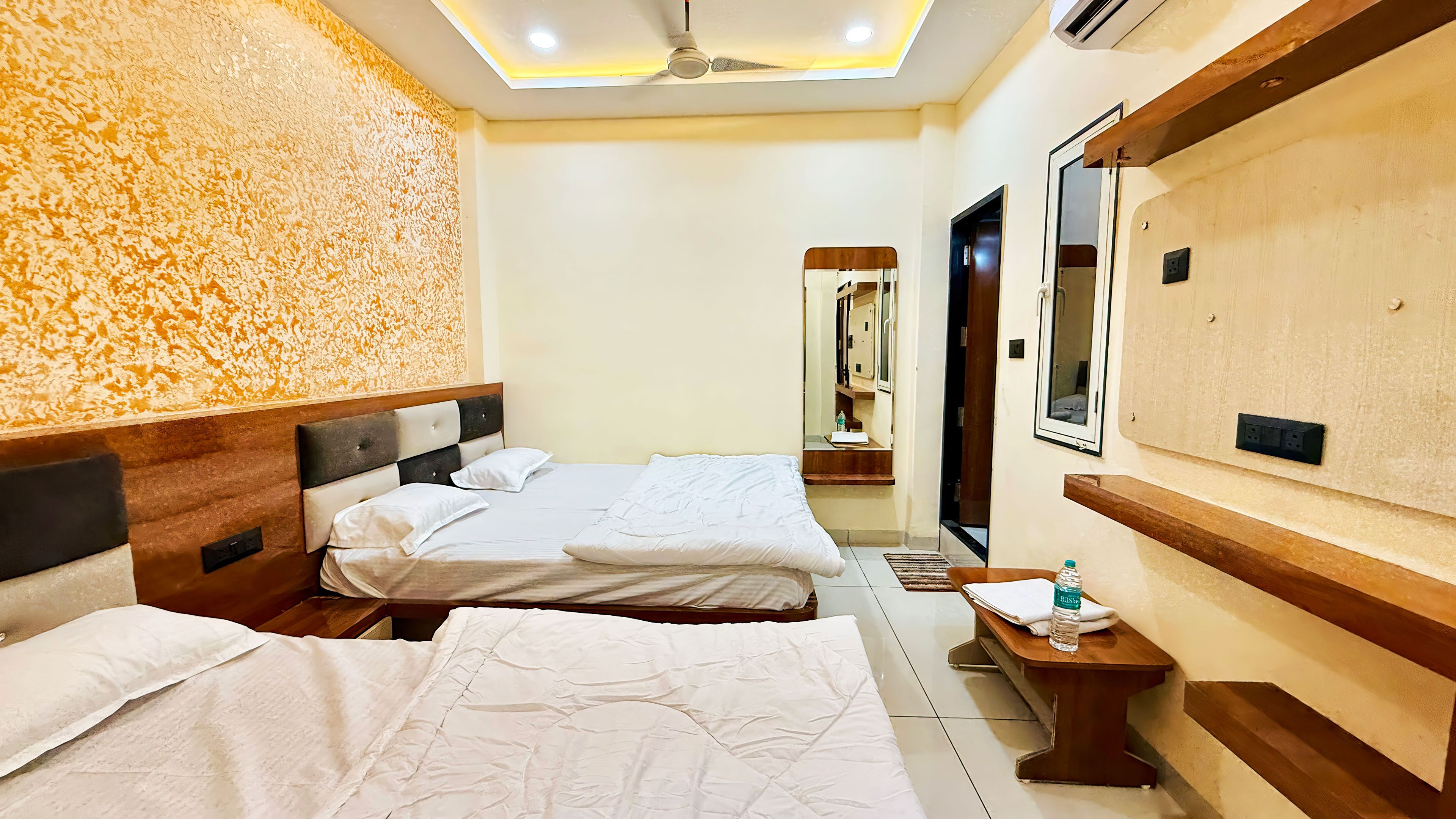 Quarto de hotel com duas camas, uma mesa de cabeceira com uma garrafa, um espelho e um ventilador de teto.