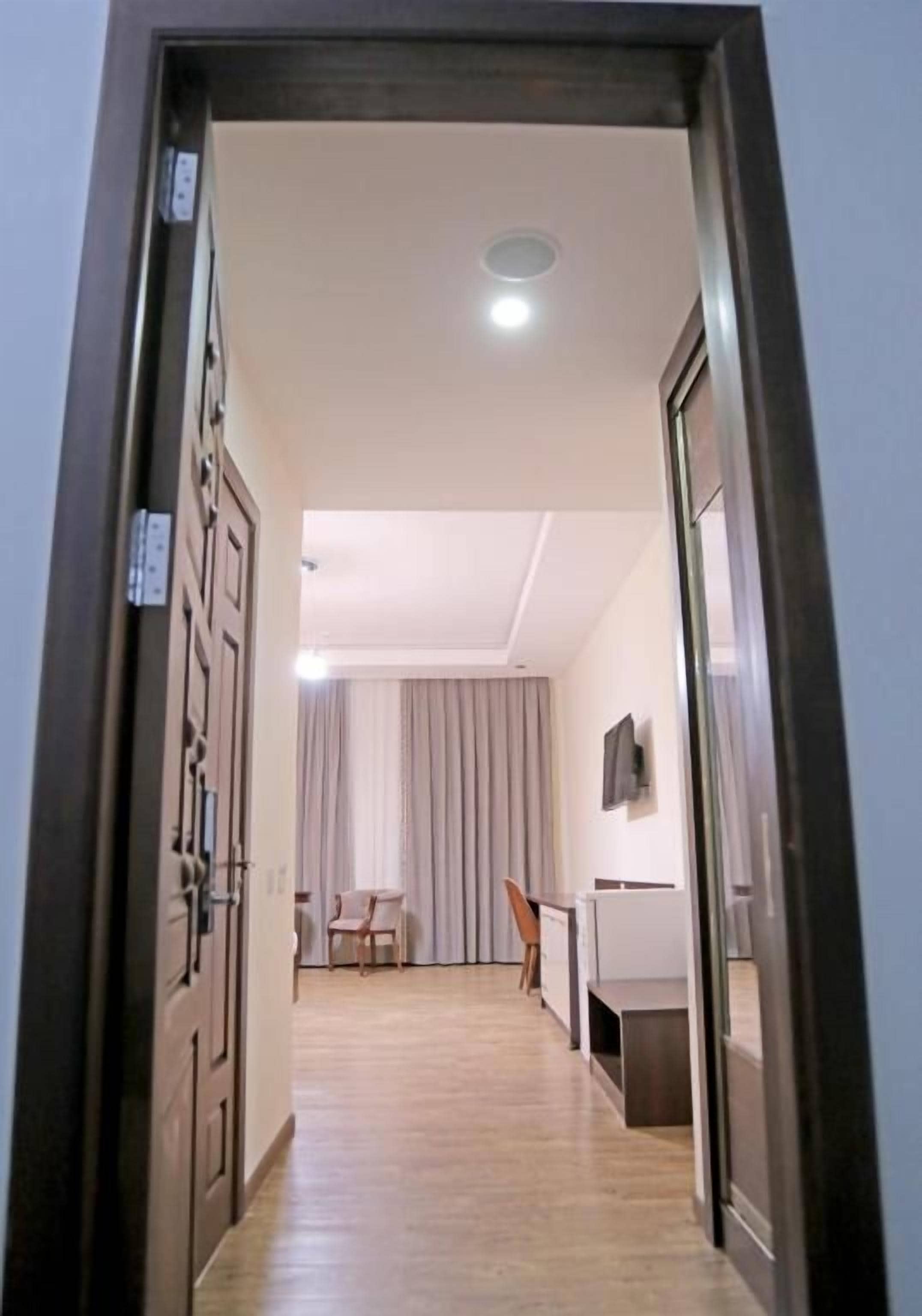 Um corredor com porta de madeira, luminária no teto, uma cadeira, uma mesa e uma cama ao fundo.