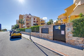 Uma rua costeira com uma fileira de carros amarelos estacionados ao longo da calçada, um muro de pedra com um portão identificado como 'P1', e edifícios altos com sacadas, sob um céu claro.