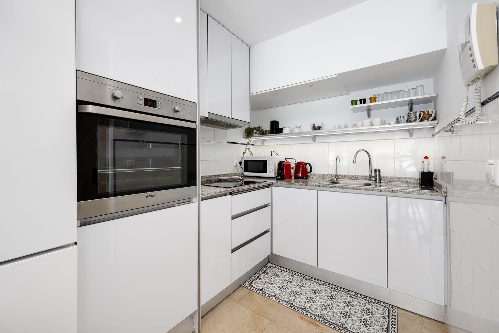 Cozinha moderna com forno embutido, micro-ondas e chaleira, armários brancos e painel de azulejos.