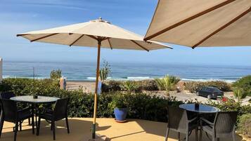 Um pátio externo com duas mesas redondas e cadeiras, um guarda-sol bege e vista para o oceano.