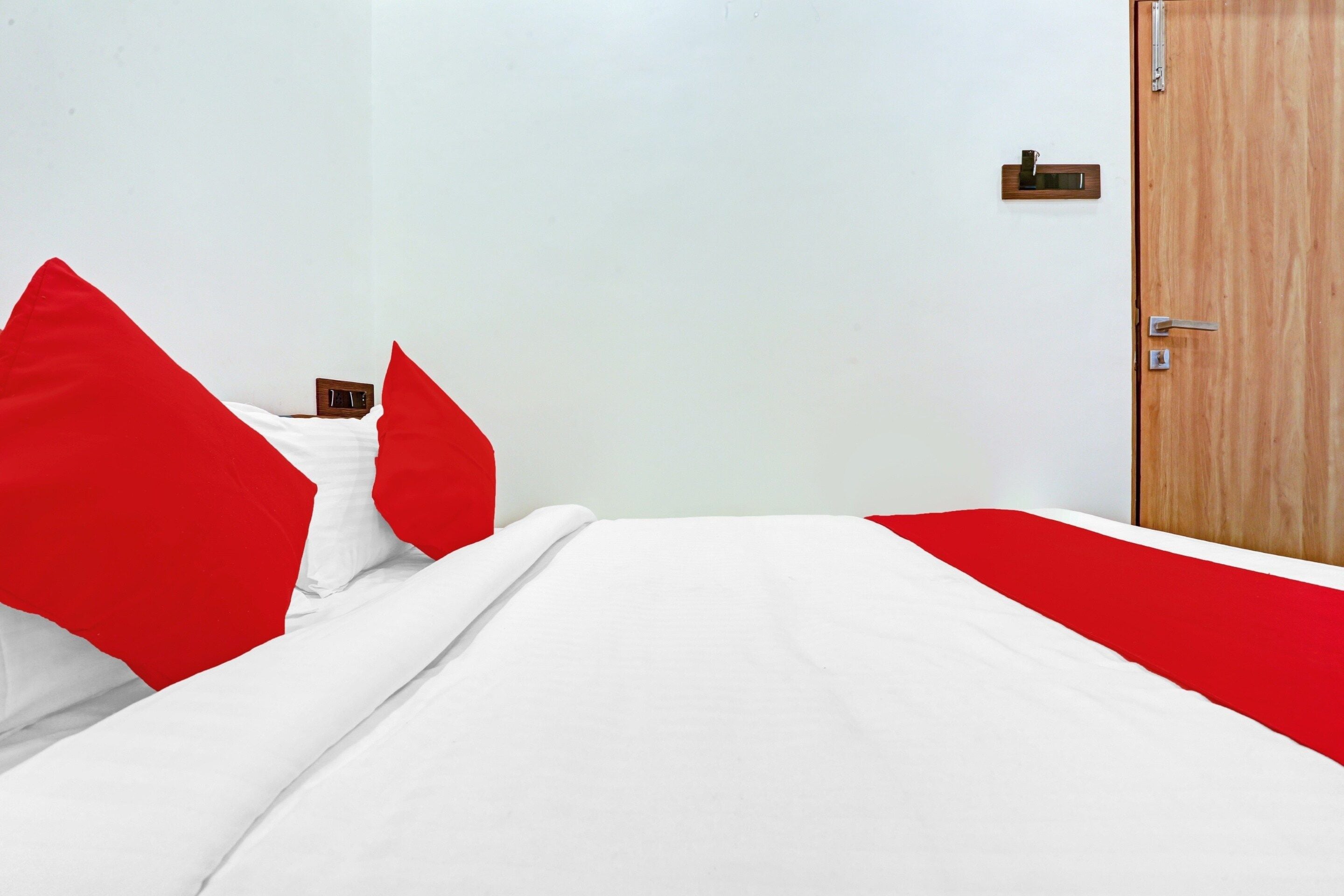 Uma cama com roupa de cama branca e almofadas vermelhas, uma mesa de cabeceira de madeira com um pequeno objeto sobre ela e uma porta de madeira com maçaneta.