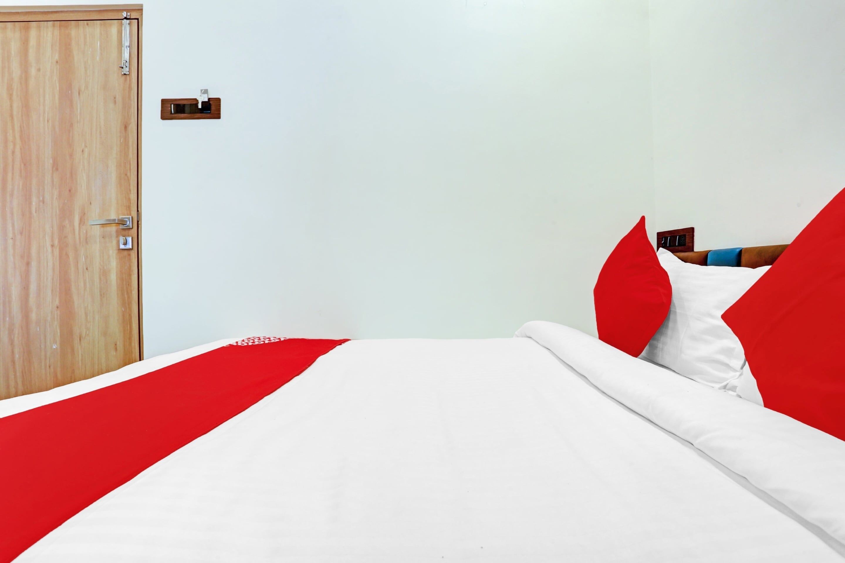 Quarto de hotel com porta de madeira, cama com roupa de cama branca e vermelha e uma pequena mesa de cabeceira com um rádio.