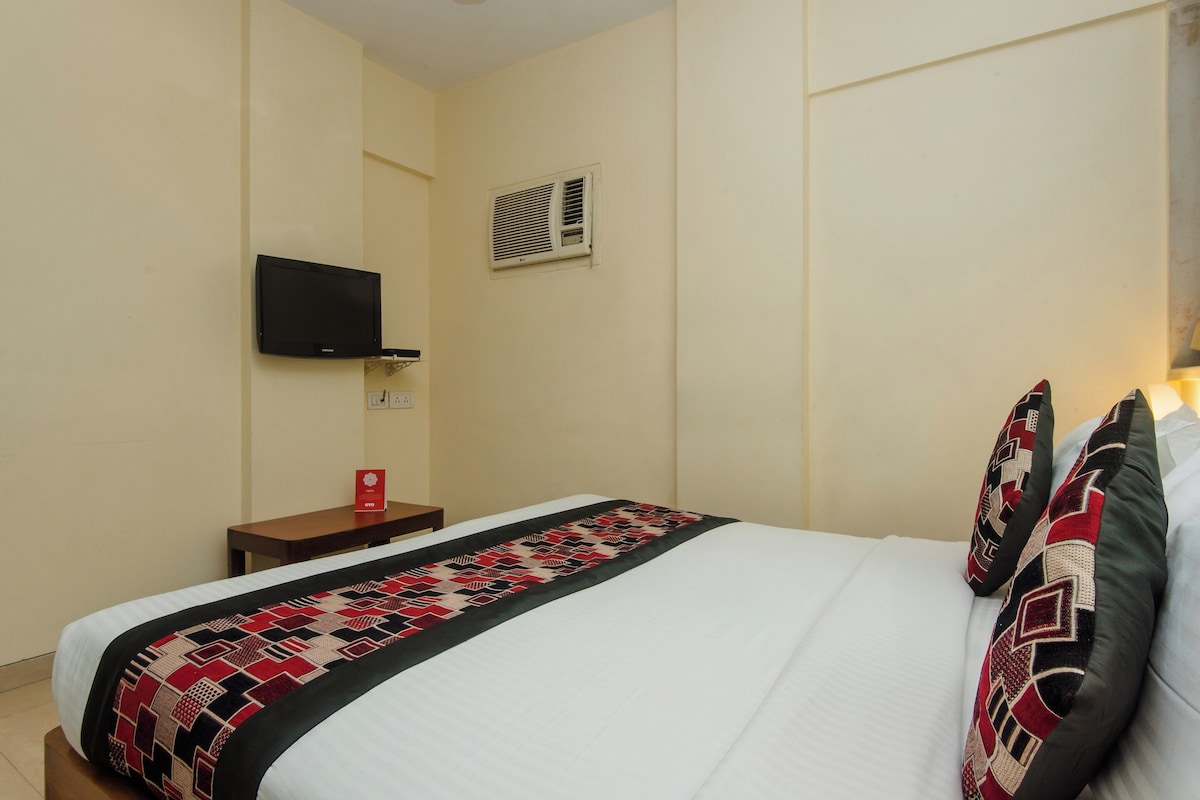 Quarto de hotel com cama, televisão fixada na parede, mesa de cabeceira e um letreiro vermelho.