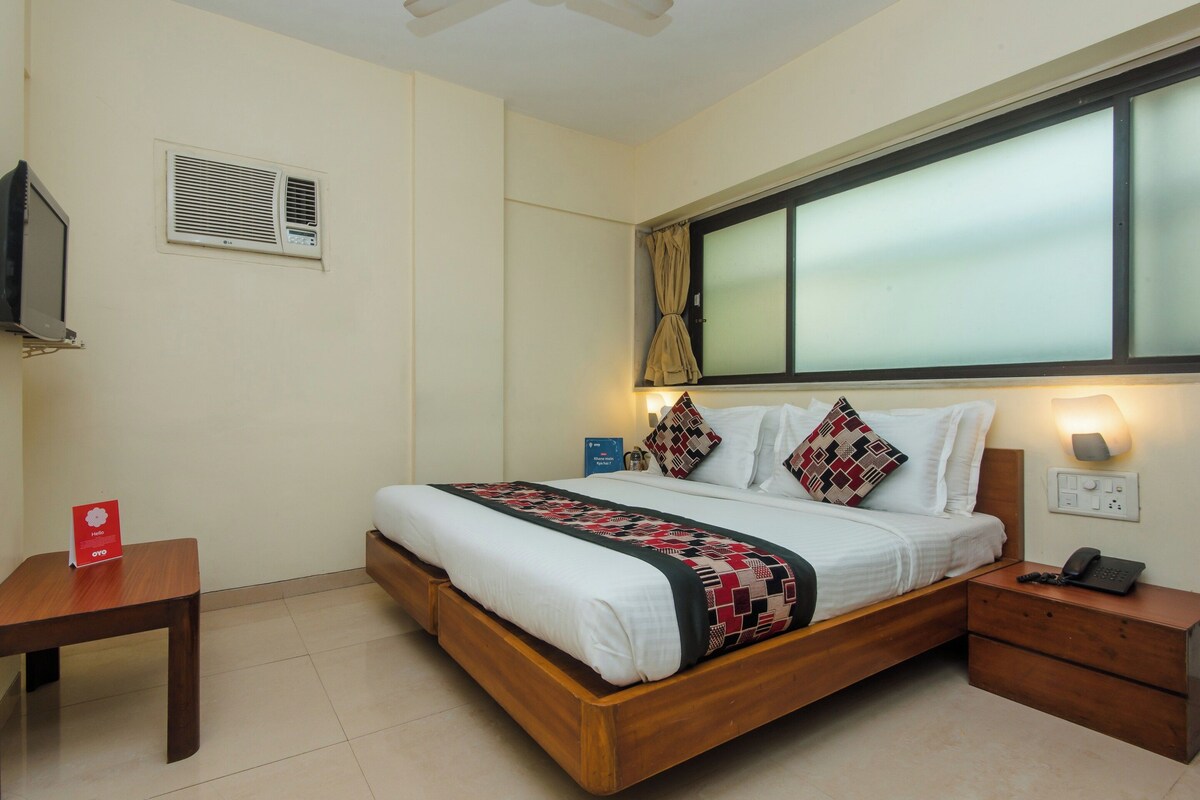 Quarto de hotel com cama de madeira, mesa de cabeceira, televisão, janela com cortinas e luminária fixada na parede.