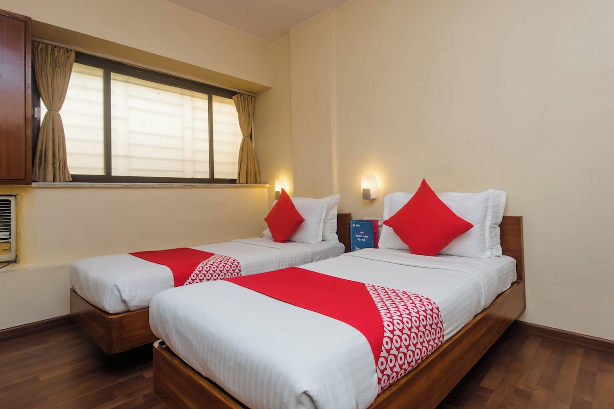 Quarto de hotel com duas camas de solteiro, cada uma com colcha vermelha e branca, uma janela com cortinas, uma luminária na parede e piso de madeira.