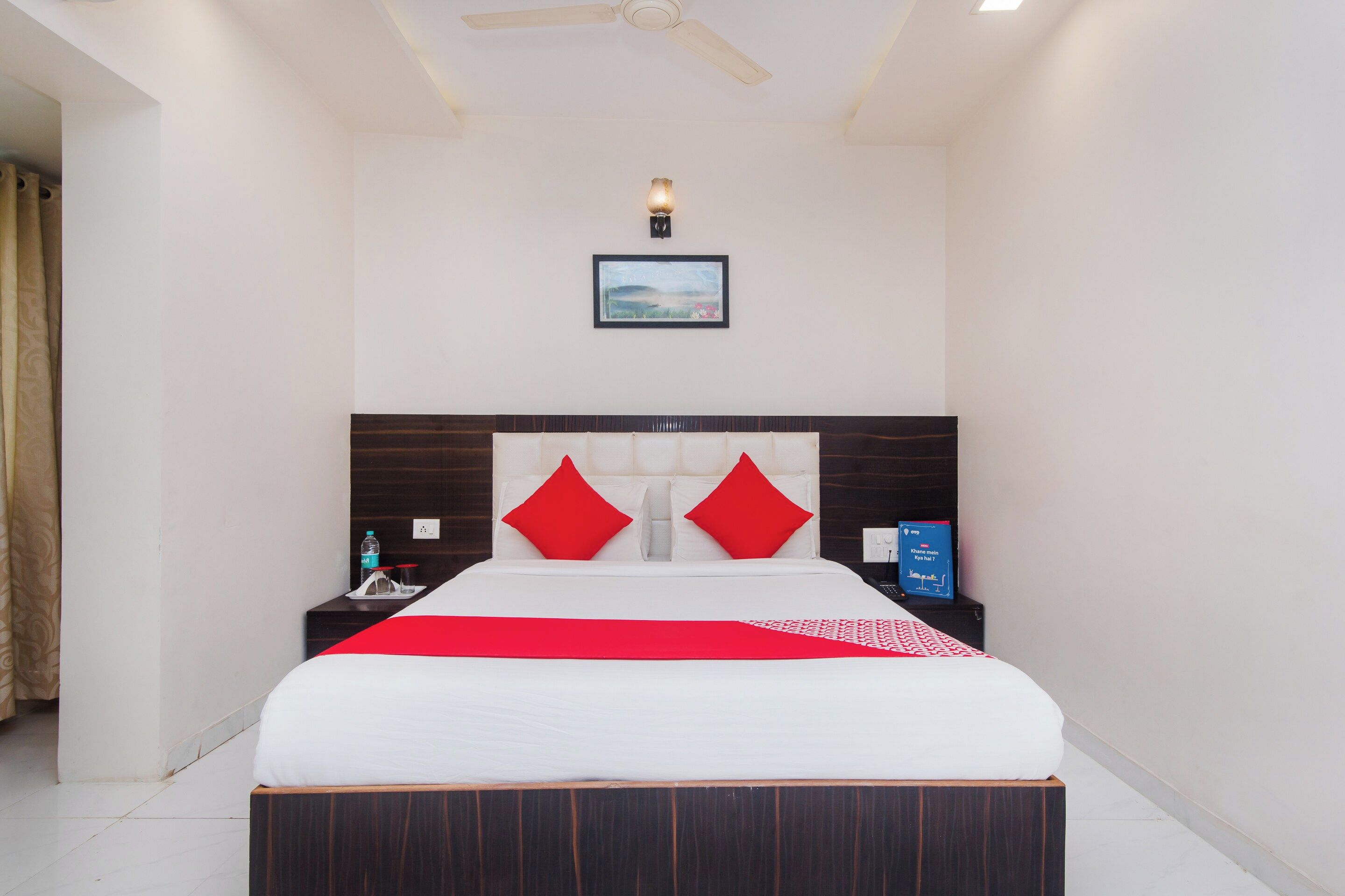 Quarto de hotel com cama, mesa de cabeceira, luminária na parede, ventilador de teto e quadro pendurado na parede.