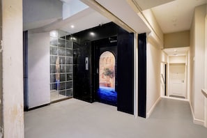 Um corredor com elevador de vidro, porta que dá para área externa e porta branca à direita.