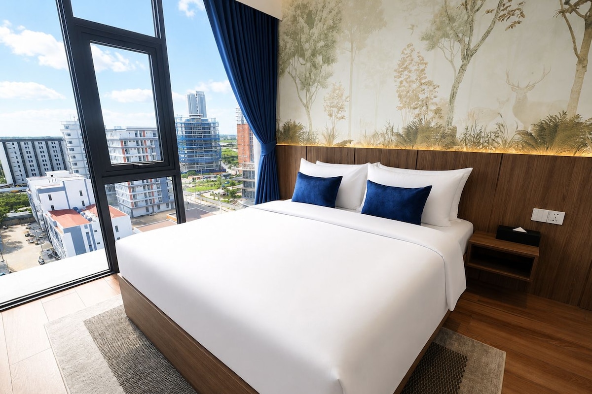Quarto de hotel com uma cama grande, uma mesa de cabeceira, uma prateleira na parede e vista da cidade por uma janela ampla.