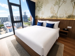 Quarto de hotel com uma cama grande, uma mesa de cabeceira, uma prateleira na parede e vista da cidade por uma janela ampla.