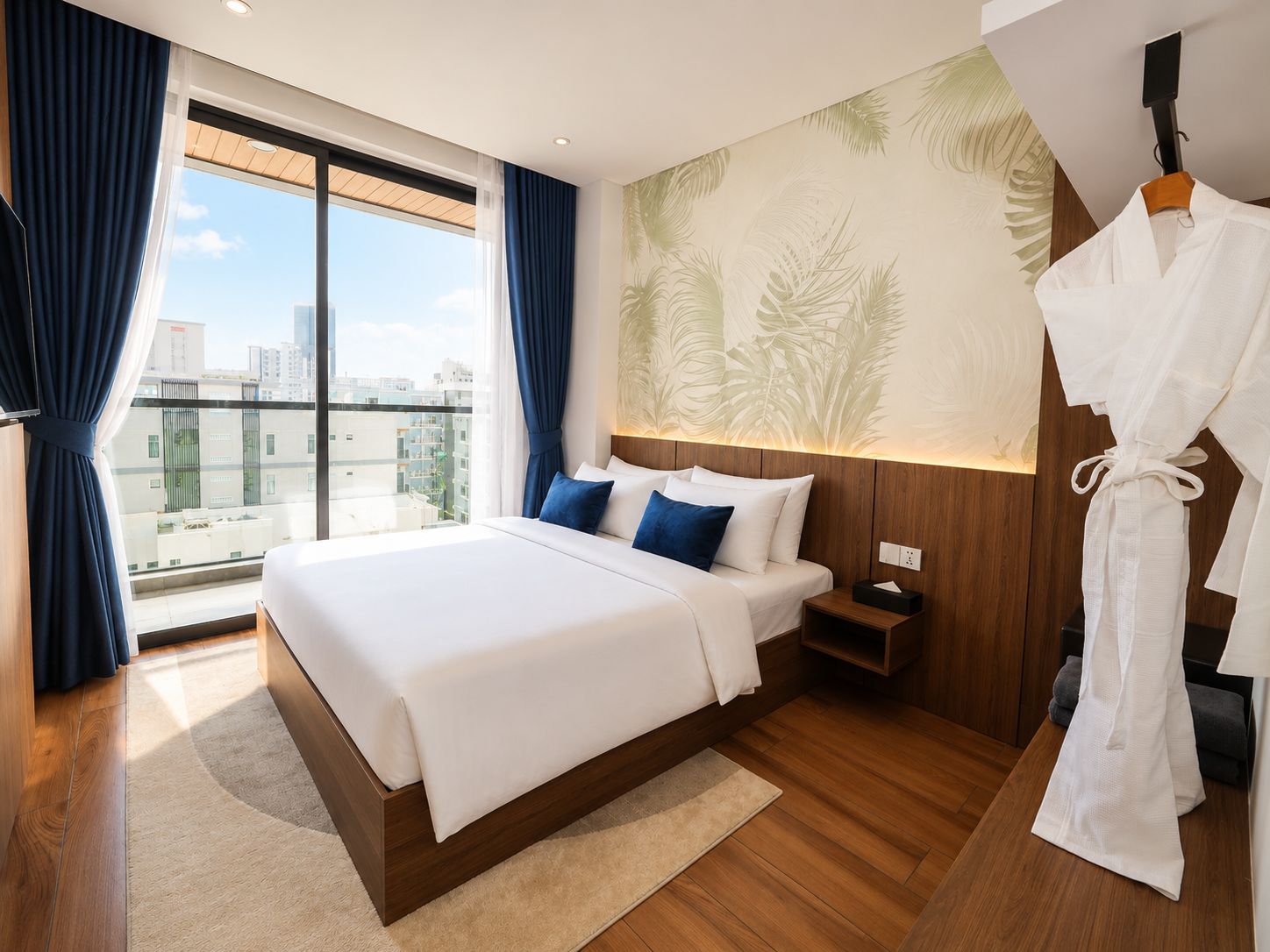 Quarto de hotel moderno com uma cama grande, cabeceira de madeira, mesa de cabeceira, roupão e vista para a cidade.