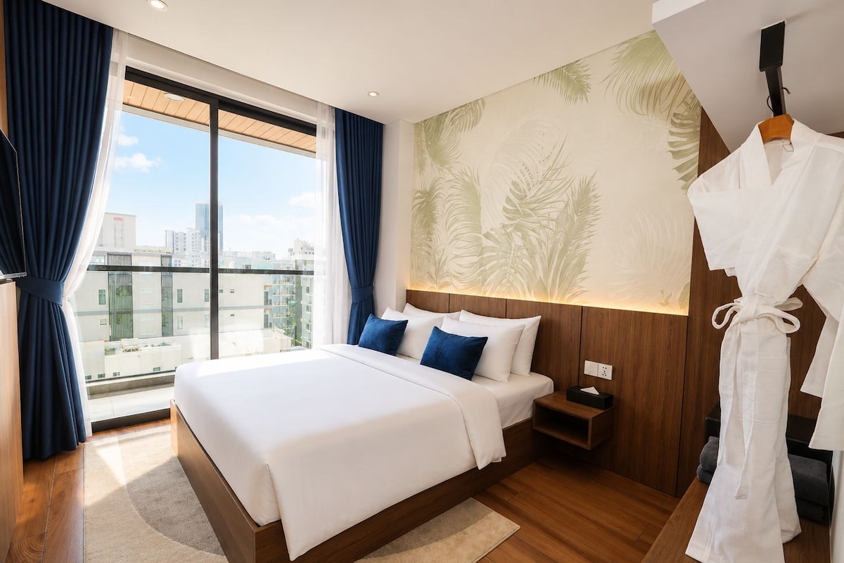 Quarto de hotel moderno com uma cama grande, cabeceira de madeira, mesa de cabeceira, roupão e vista para a cidade.