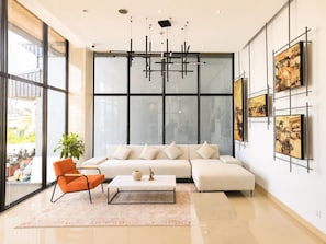 Uma sala de estar moderna com um sofá branco, uma poltrona laranja e uma mesa de centro. Há janelas amplas e obras de arte nas paredes.