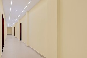 Um corredor comprido e estreito com iluminação contínua no teto, paredes lisas e portas com molduras escuras.