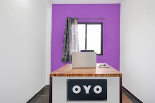 Quarto com parede roxa, janela com cortina, escrivaninha de madeira com laptop e placa com a inscrição 'OYO'.