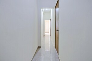Um corredor com uma porta de madeira à direita, que dá para um quarto com piso de azulejos.