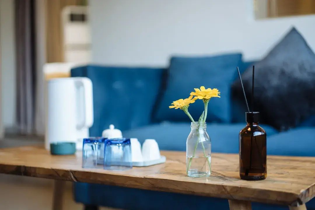 Uma mesa de madeira com um vaso de vidro transparente contendo flores amarelas, uma garrafa marrom e um objeto azul.