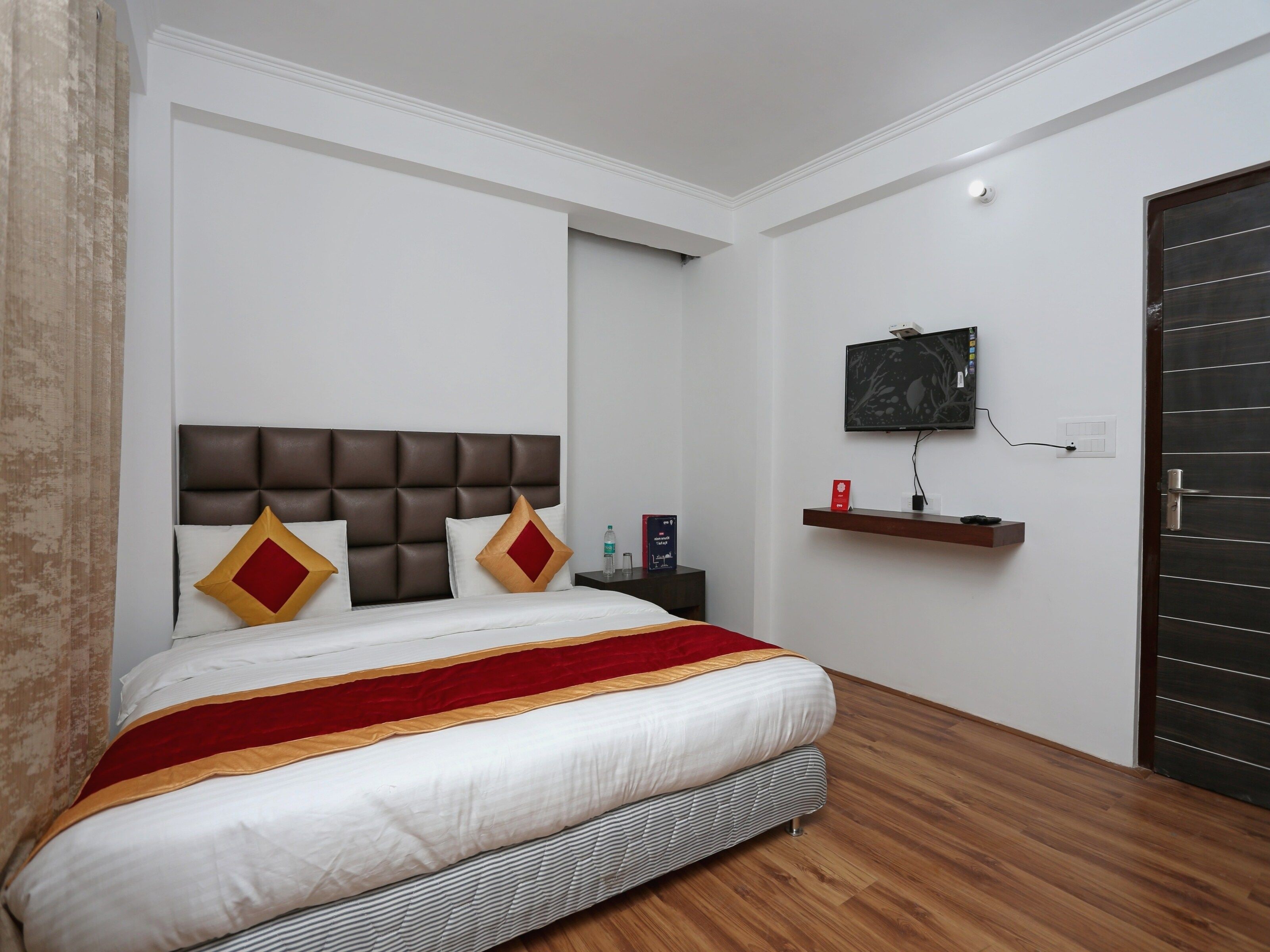 Quarto de hotel com cama, televisão na parede, piso de madeira e uma porta.