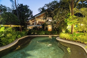 Piscina com design curvo, rodeada por vegetação exuberante e um edifício com vários níveis e iluminação externa.