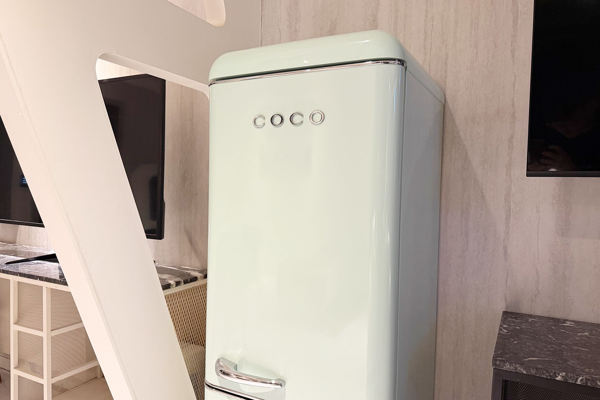 Geladeira de cor clara, estilo vintage, com puxador e painel de controle cromados, em cozinha com parede de madeira e bancada preta.
