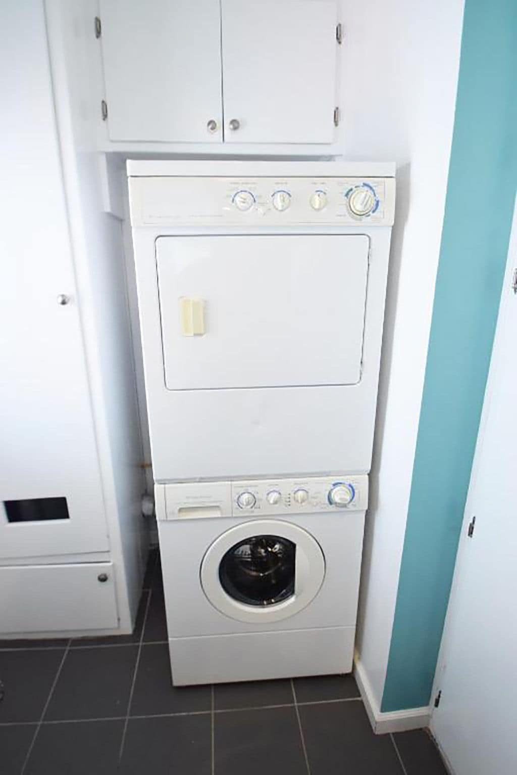 Uma lavanderia compacta e clara, com máquina de lavar e secadora embutidas, um pequeno armário acima da secadora e piso revestido de azulejos.