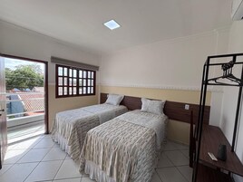 Quarto de hotel com duas camas, janela com vista para o exterior, luminária no teto e uma mesa pequena com um alto-falante.