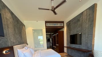 Quarto de hotel moderno com cama, mesa de cabeceira, um pequeno espaço de estar, ventoinha no teto e TV fixada na parede.