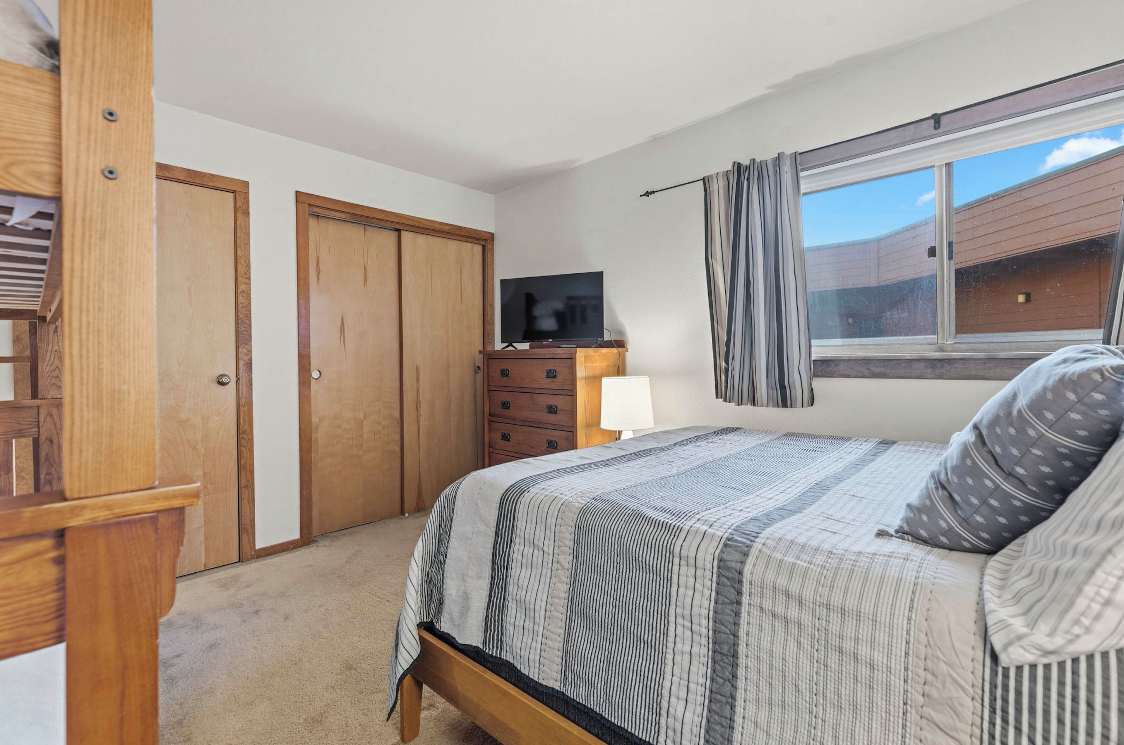 Quarto com cama, televisão sobre uma cômoda de madeira, janela com cortinas e porta.