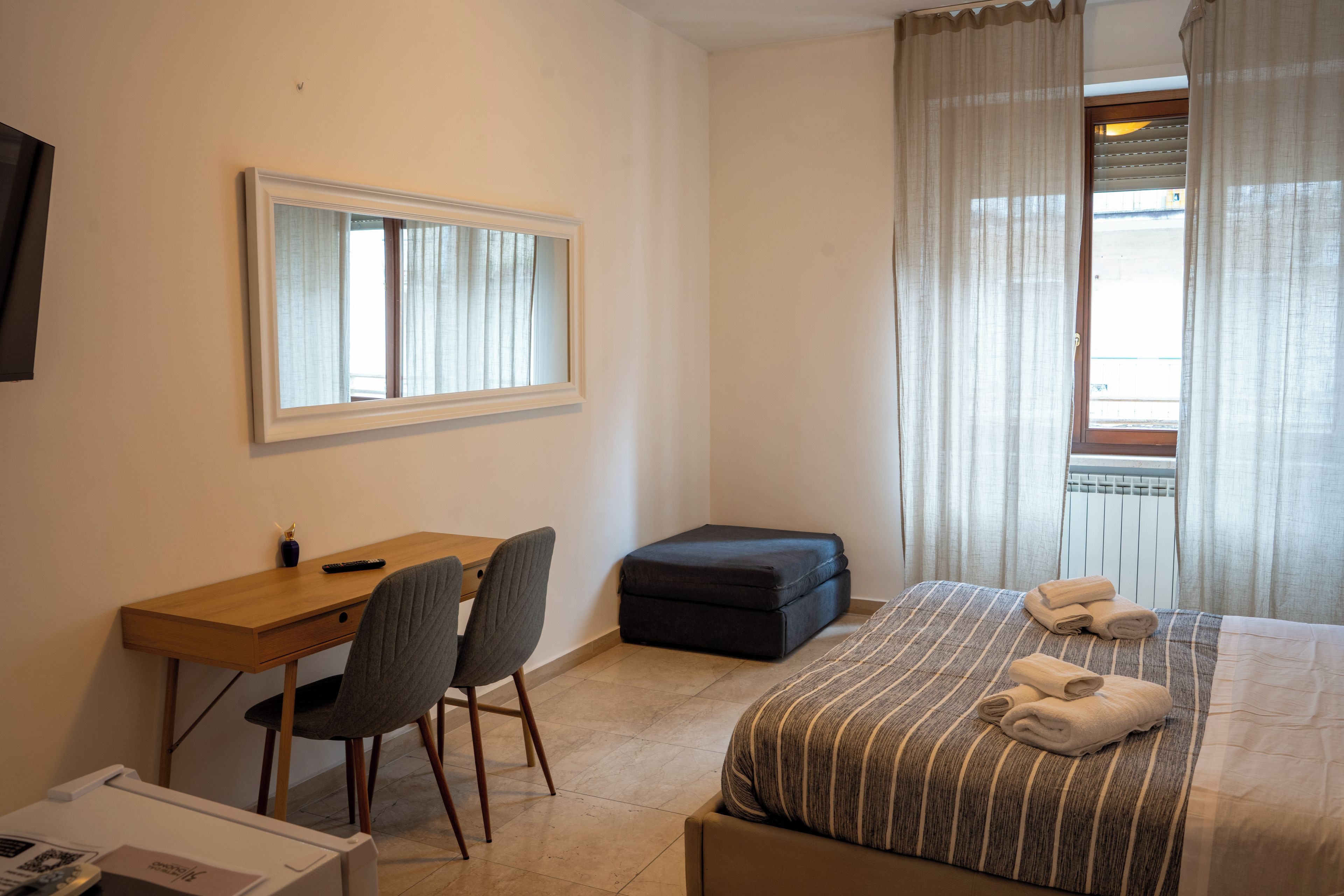 Quarto de hotel com cama, escrivaninha com cadeira, um pequeno banquinho, televisão e janela com cortinas.