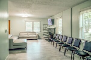 Quarto com sofá bege, fileira de cadeiras pretas, televisão na parede e janela com persianas.