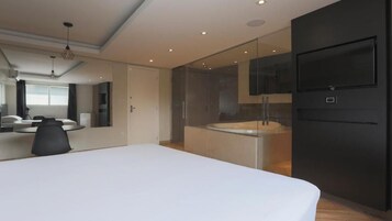 Quarto de hotel moderno com uma cama grande, banheiro com paredes de vidro e televisão de tela plana na parede.