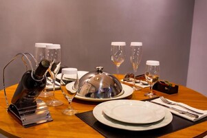 Mesa posta com pratos, copos, garrafa de vinho, abridor de garrafa e um prato com comida coberta.