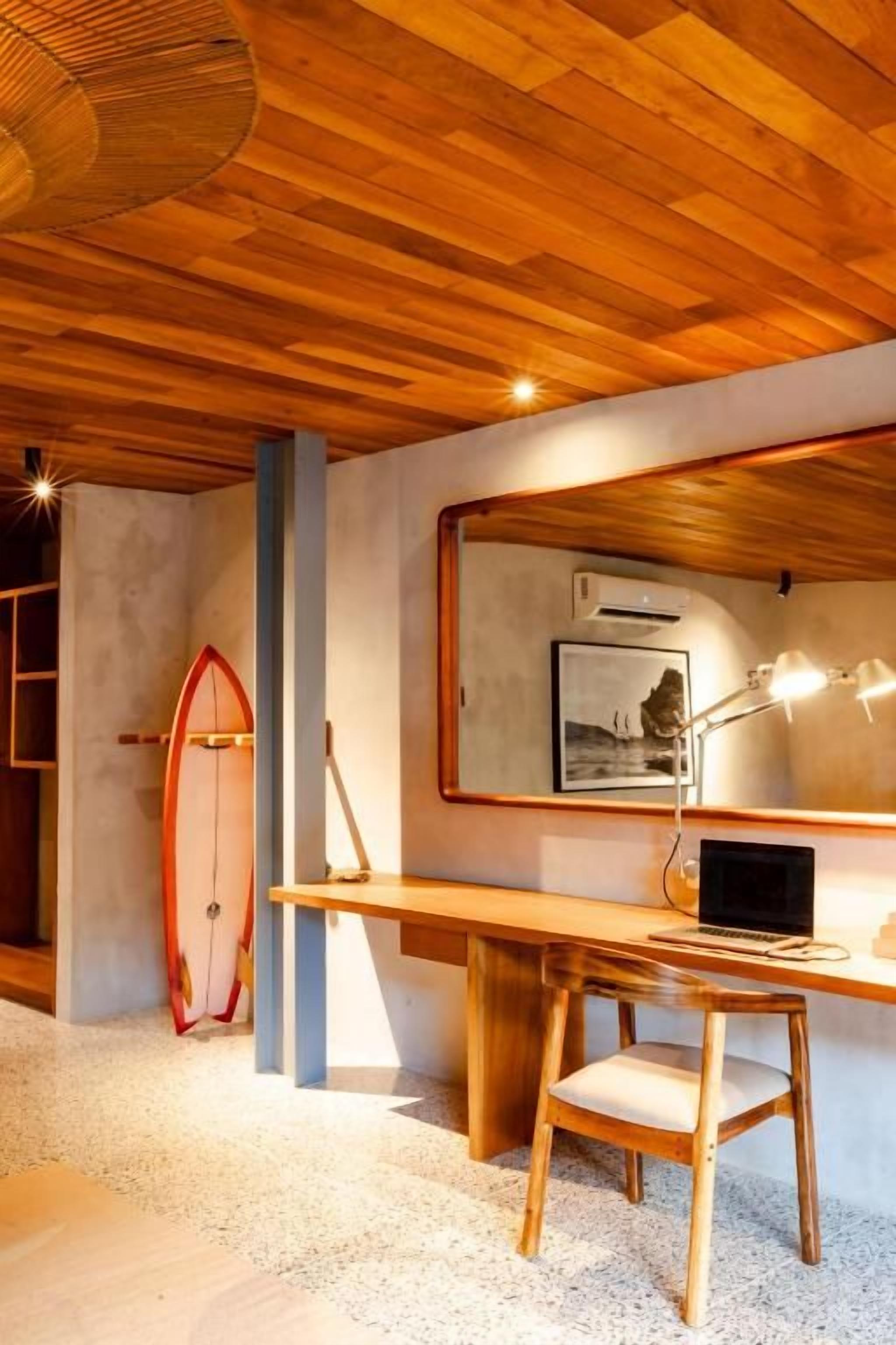 Quarto com escrivaninha de madeira, cadeira, prancha de surf e uma moldura na parede.