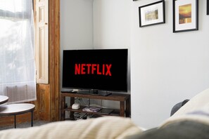 Quarto com televisão exibindo 'NETFLIX' na tela, uma pequena mesa de madeira, uma cadeira e uma janela com cortinas leves.