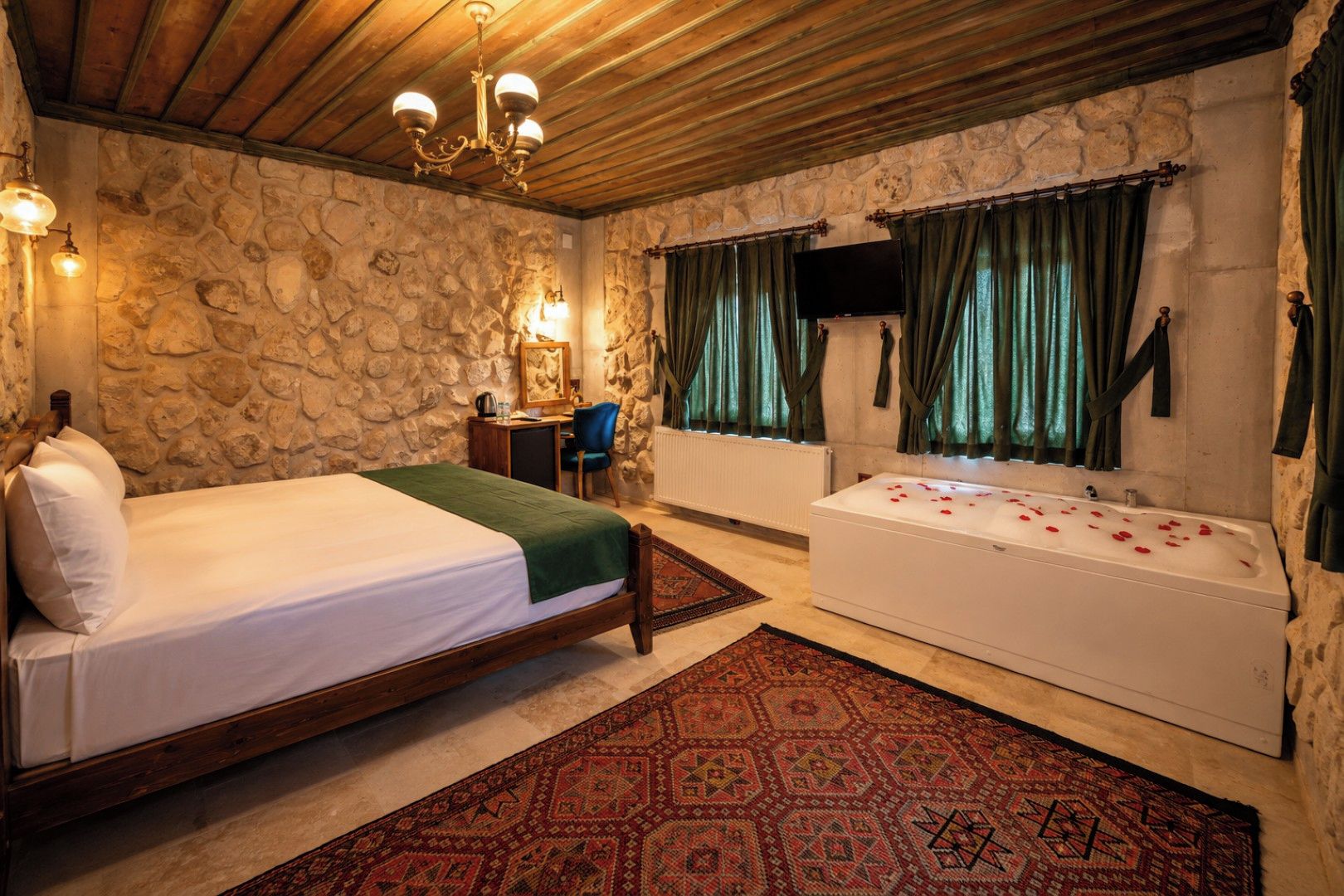 Quarto com cama, escrivaninha, cadeira, banheira com pétalas de rosa e parede de pedra.