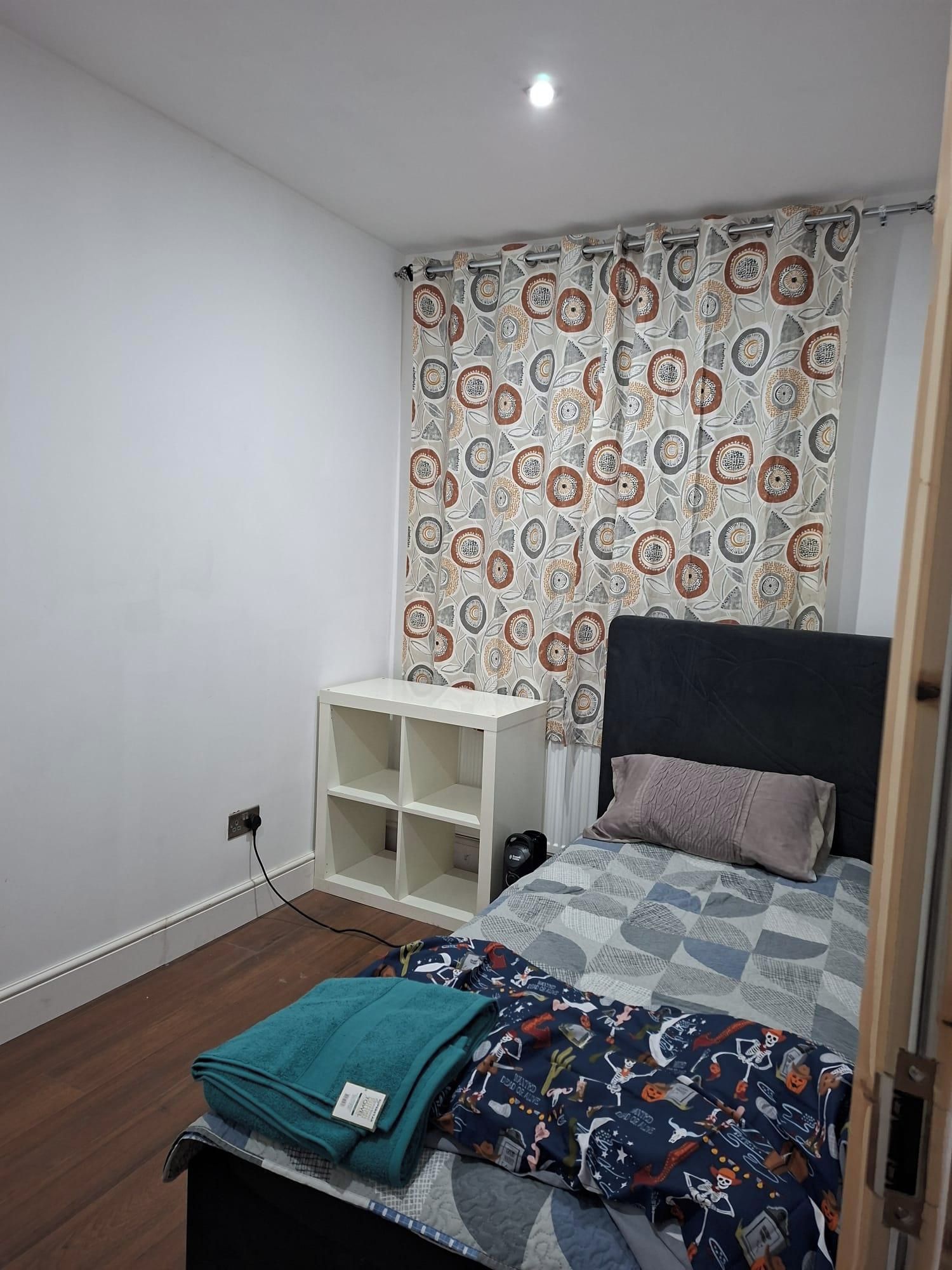 Quarto com cama, mesa de cabeceira, câmara e cortina com padrão geométrico.