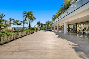 Um corredor amplo ao ar livre, com palmeiras, piso de azulejos e edifícios modernos ao fundo.
