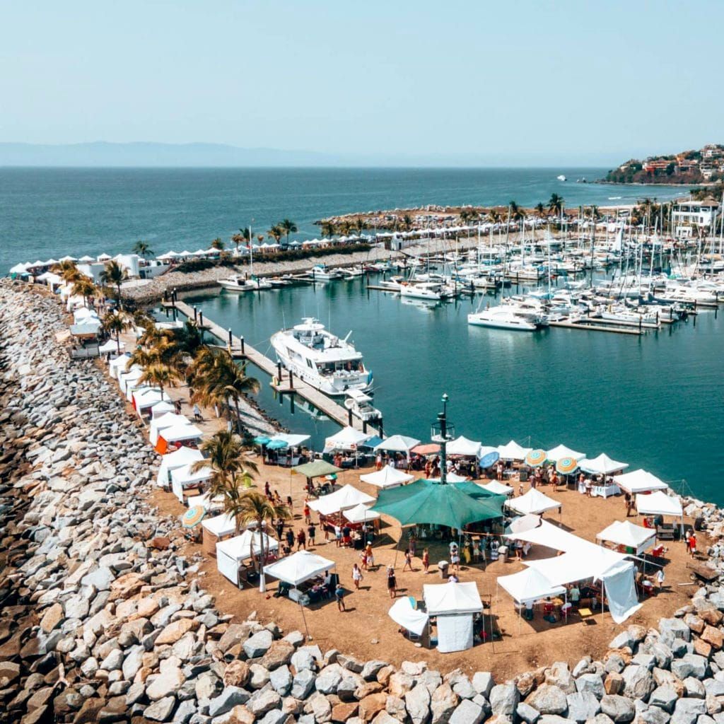 Uma marina com barcos atracados, uma praia arenosa com guarda-sóis e terreno rochoso.