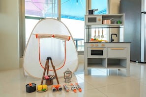 Quarto com uma janela grande, escrivaninha branca com gavetas, prateleira com utensílios de cozinha e uma estrutura tipo tenda com lanterna e ferramentas.