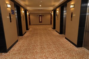 Corredor de hotel com piso coberto de carpete, portas de elevador e luminárias fixadas na parede.