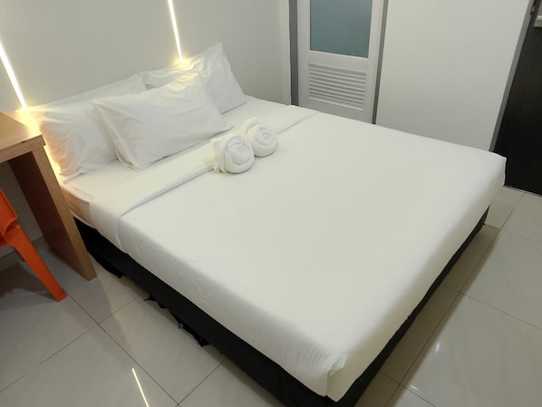 Cama bem arrumada com lençóis brancos e duas toalhas enroladas, mesa de cabeceira de madeira com abajur, e janela com persianas.