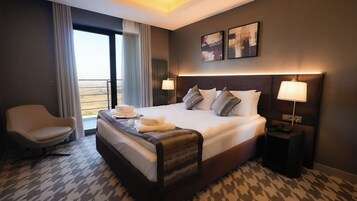Quarto de hotel com uma cama grande, uma cadeira, mesa de cabeceira com abajur e uma obra de arte na parede.